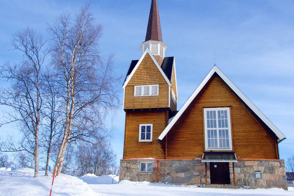 Uma igreja de madeira com um pináculo, rodeada por neve e árvores sem folhas.