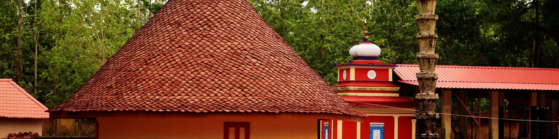 Um edifício tradicional com telhado de telhas vermelhas e uma pequena porta, rodeado por vegetação e palmeiras.