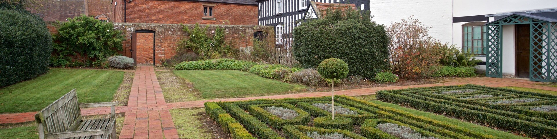 Um jardim com um caminho de tijolos, um banco de madeira, sebes bem aparadas e casas em estilo Tudor.