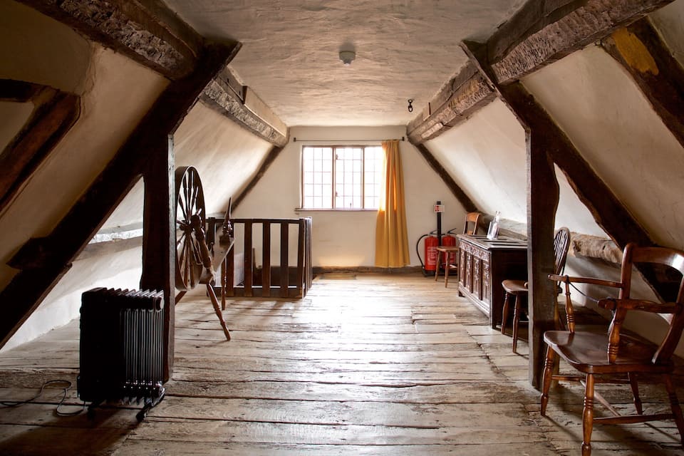 Quarto rústico no sótão, com vigas de madeira, radiador, roda de fiar e janela com cortinas.