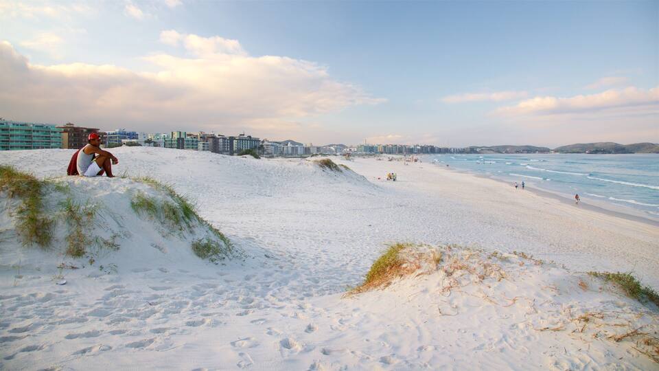 Uma praia arenosa com pegadas, uma pessoa vestida de vermelho e edifícios ao fundo.
