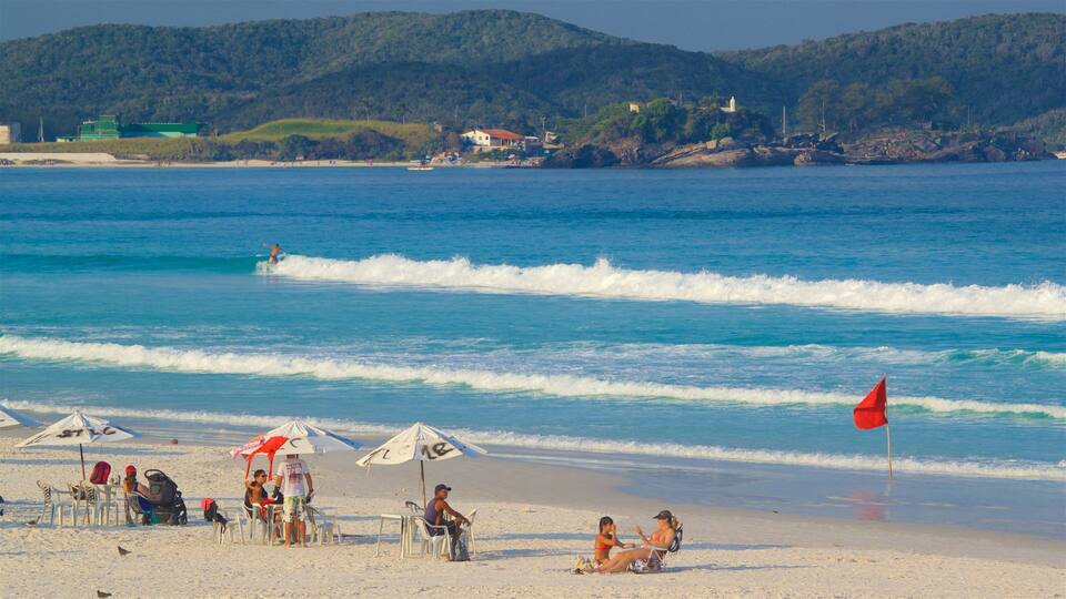 Uma praia com pessoas relaxando sob guarda-sóis, uma bandeira vermelha indicando proibição de banho e ondas se aproximando da costa.