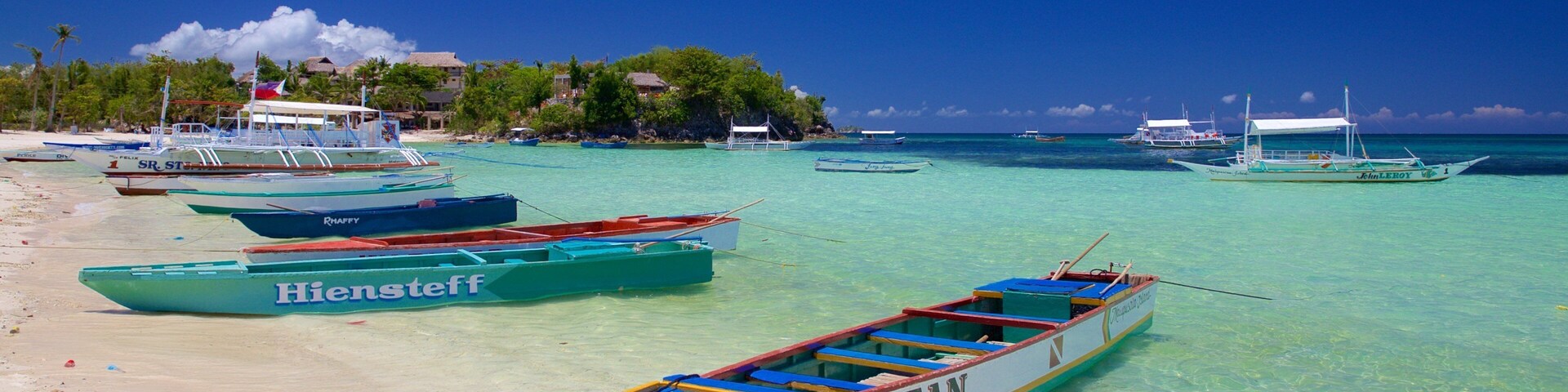 Uma praia com vários barcos coloridos ancorados perto da costa, água turquesa cristalina e um cenário de vegetação exuberante e edifícios.