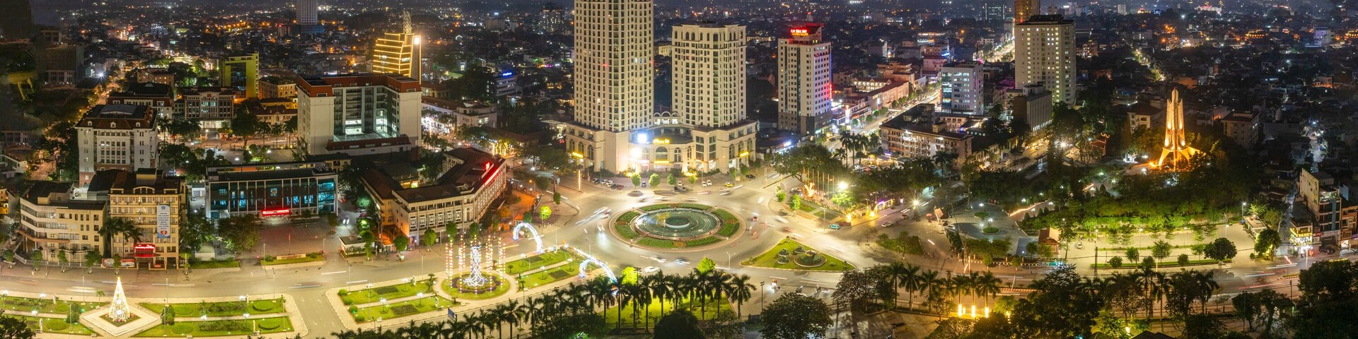 Paisagem urbana ao anoitecer, com edifícios iluminados, uma rotatória central e uma cadeia de montanhas ao longe.