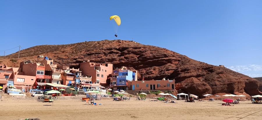 Uma vila costeira com edifícios coloridos, uma praia arenosa e uma pessoa a parapontar ao fundo.