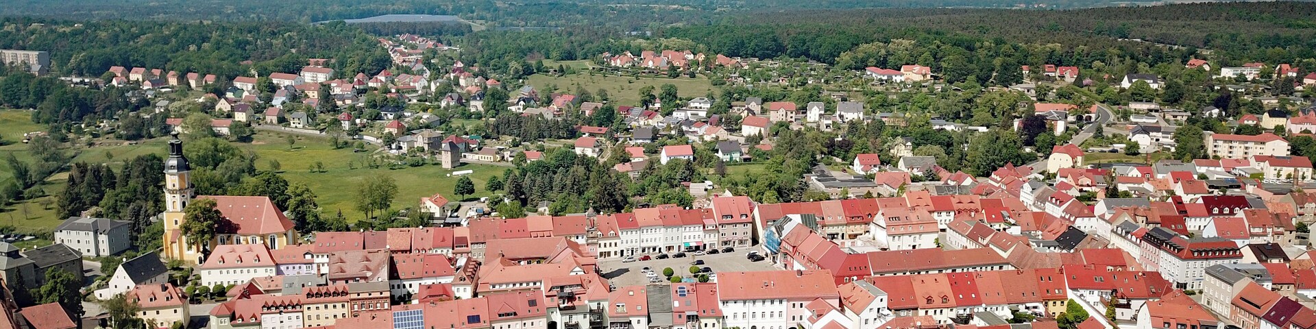 Uma cidade com edifícios de telhados vermelhos, uma torre de igreja imponente e um lago ao fundo.