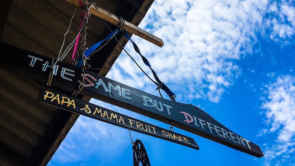 Um letreiro com o texto 'THE SAME BUT DIFFERENT' e 'PAPA & MAMA FRUIT SHAKE' contra um céu azul com nuvens.