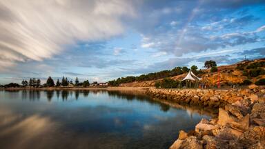 Um lago calmo com uma tenda branca numa margem rochosa, árvores ao fundo e um arco-íris no céu.