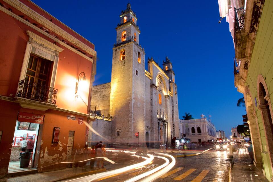 Um edifício histórico com torre do relógio, uma rua com rastros de luz de veículos em movimento e um edifício vermelho com uma placa.