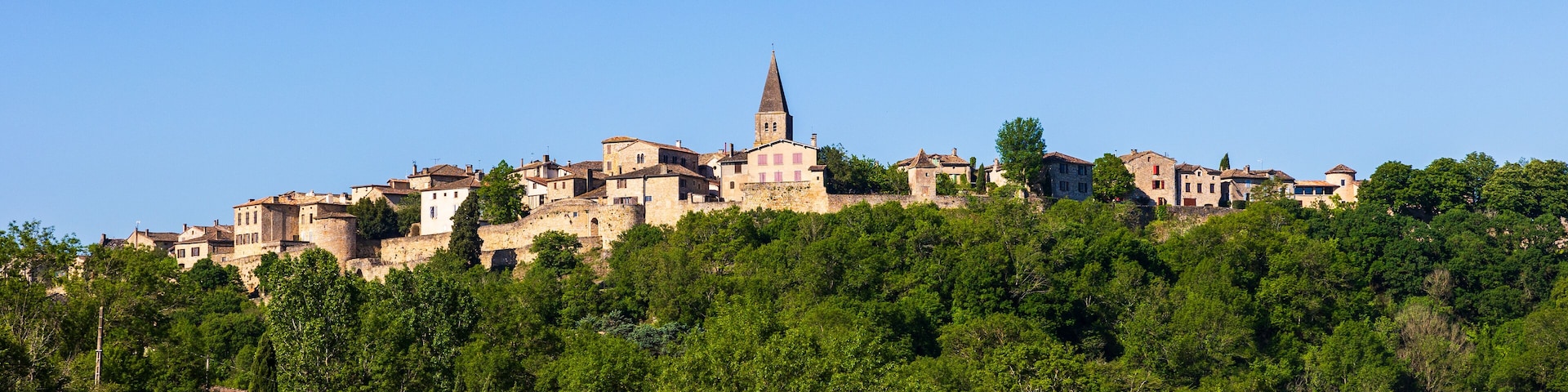 Uma cidade histórica com uma torre de igreja imponente numa colina, rodeada por vegetação exuberante e campos.