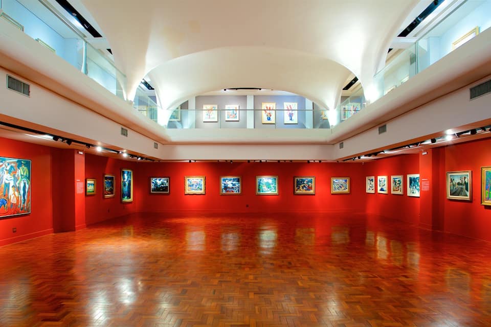 Uma galeria de arte com piso de madeira e paredes vermelhas, exibindo diversas pinturas.