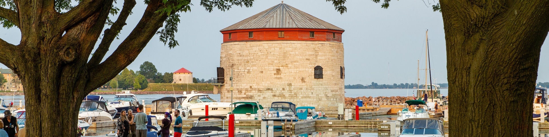 Uma torre histórica com telhado vermelho, rodeada por barcos e árvores numa marina.