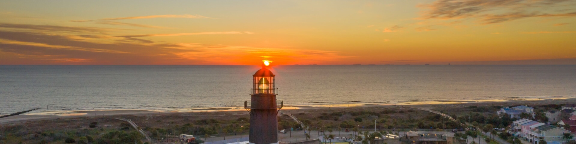 Uma vila costeira ao pôr do sol, com um farol imponente, praia e área residencial.