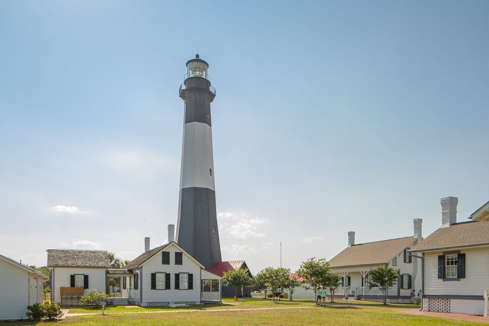 Um farol alto com uma torre listrada de branco e preto, rodeado por edifícios históricos brancos com persianas escuras.