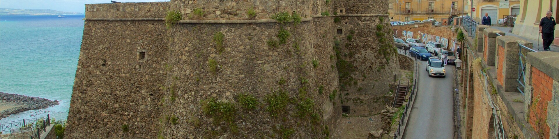 Fortificação de pedra com vista para uma estrada costeira, carros estacionados e uma placa de pare.