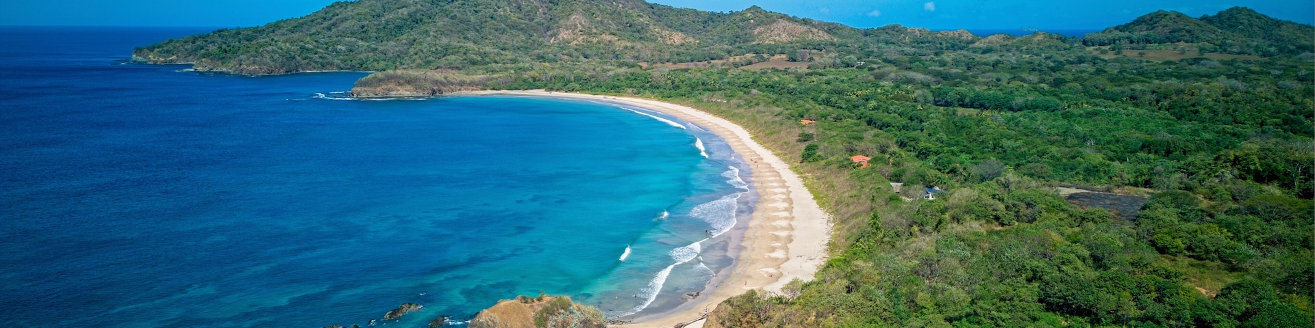 Paisagem costeira com praia arenosa, águas azuis cristalinas e colinas verdes ao fundo.