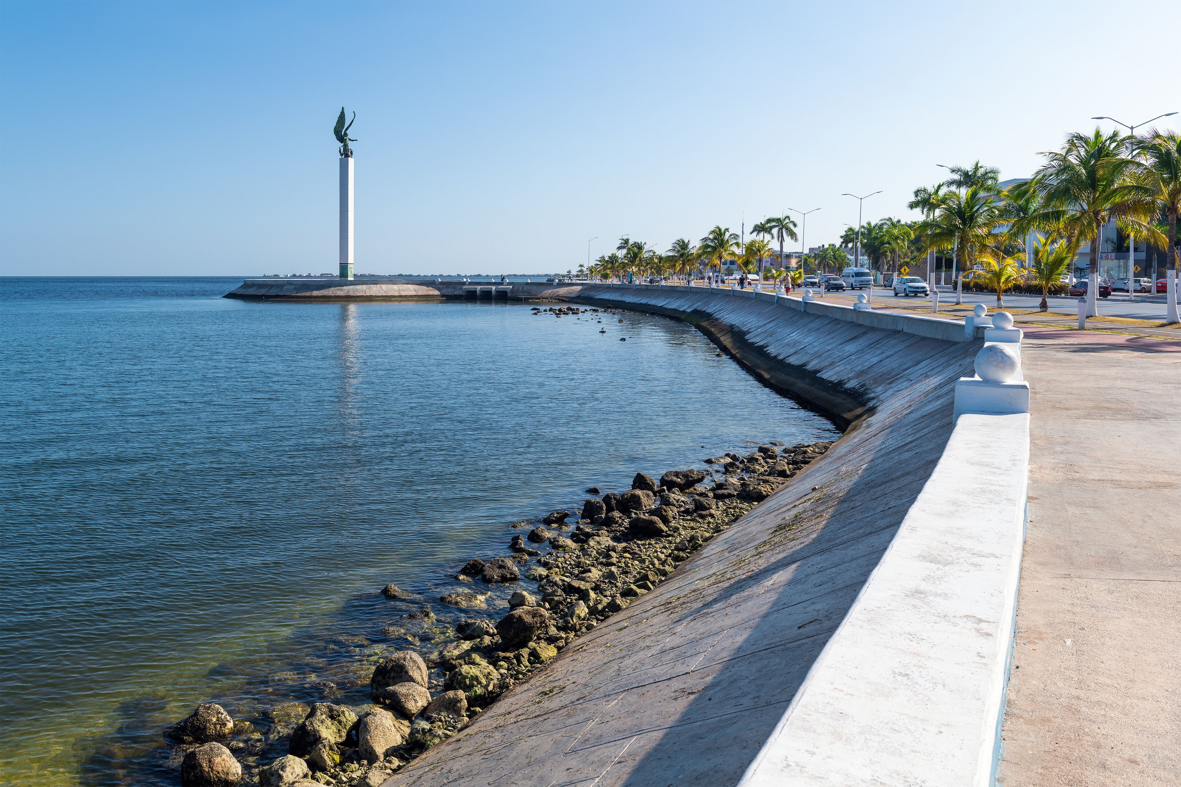 Um calçadão costeiro com um farol, palmeiras e um mar tranquilo.