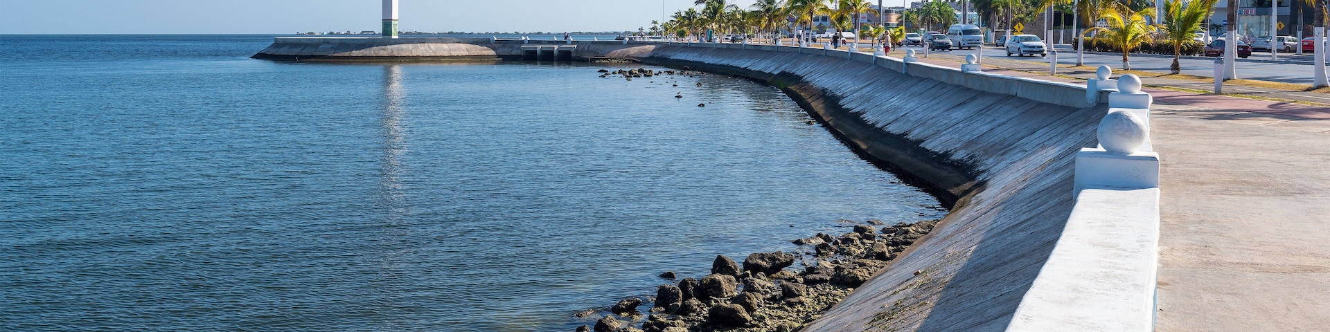 Um calçadão costeiro com um farol, palmeiras e um mar tranquilo.