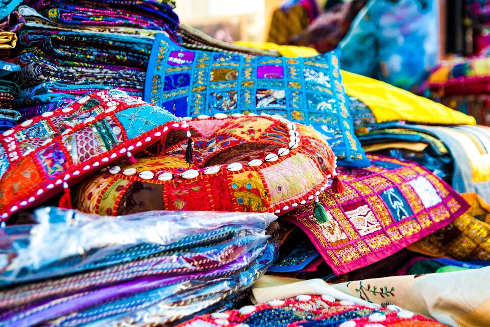 Diversidade de tecidos e estampas coloridas, incluindo almofadas e cobertores bordados, expostos em um mercado.