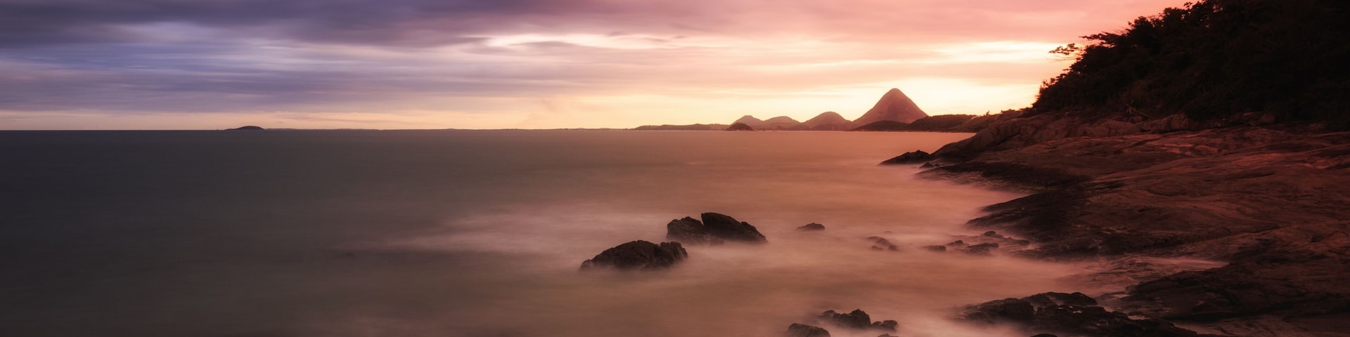 Paisagem costeira com montanha ao longe, linha de costa rochosa e mar nevoento.