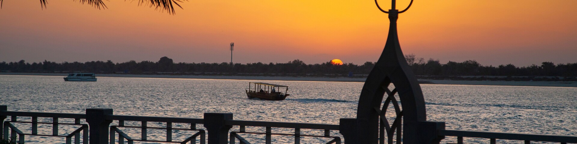 Um pôr do sol sobre um rio, com grade de ponte, um barco e um poste de luz.