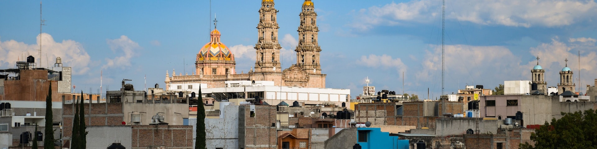 Paisagem urbana com uma igreja em destaque, apresentando duas torres altas e uma cúpula dourada, cercada por edifícios bem próximos.