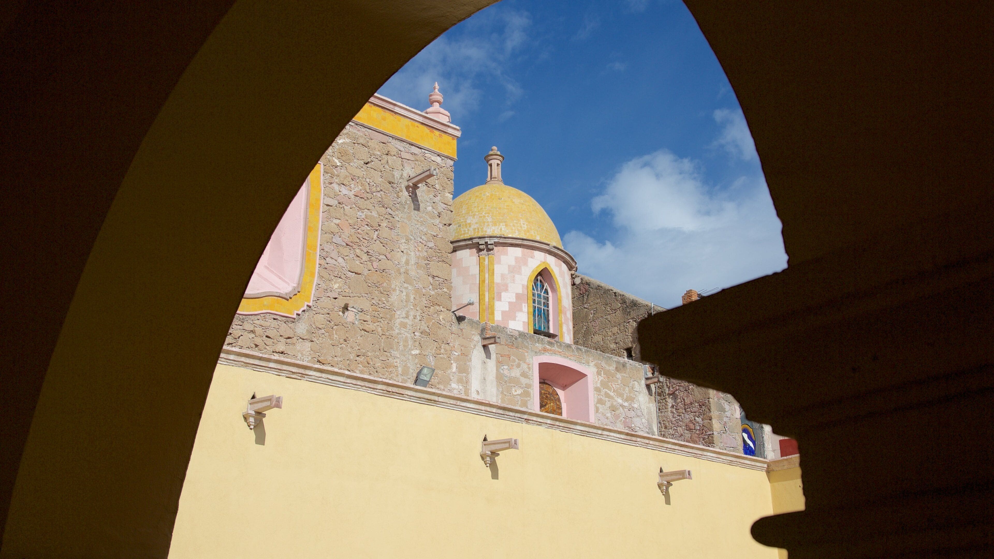 Uma igreja com cúpula amarela e paredes cor-de-rosa, vista através de um arco.