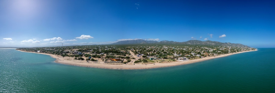 Vista aérea de uma cidade costeira com praia, casas e montanhas ao fundo.