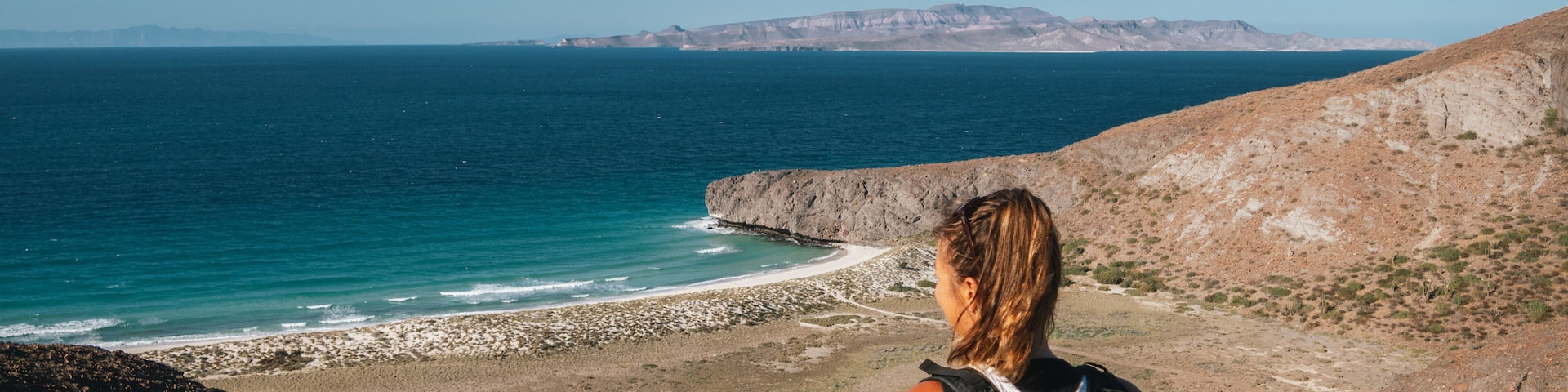 Uma pessoa em uma encosta rochosa, com vista para a paisagem costeira, o oceano e montanhas ao longe.