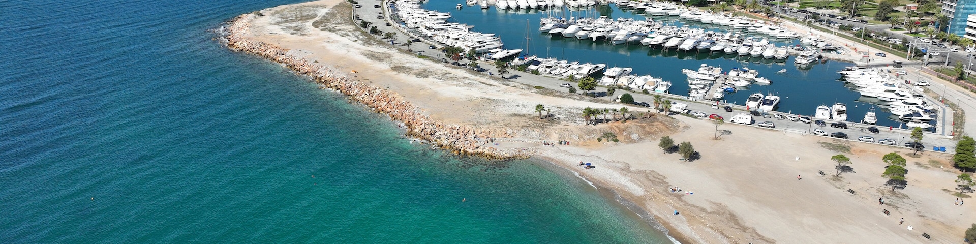 Uma marina costeira com barcos, uma praia arenosa e um horizonte urbano ao fundo.