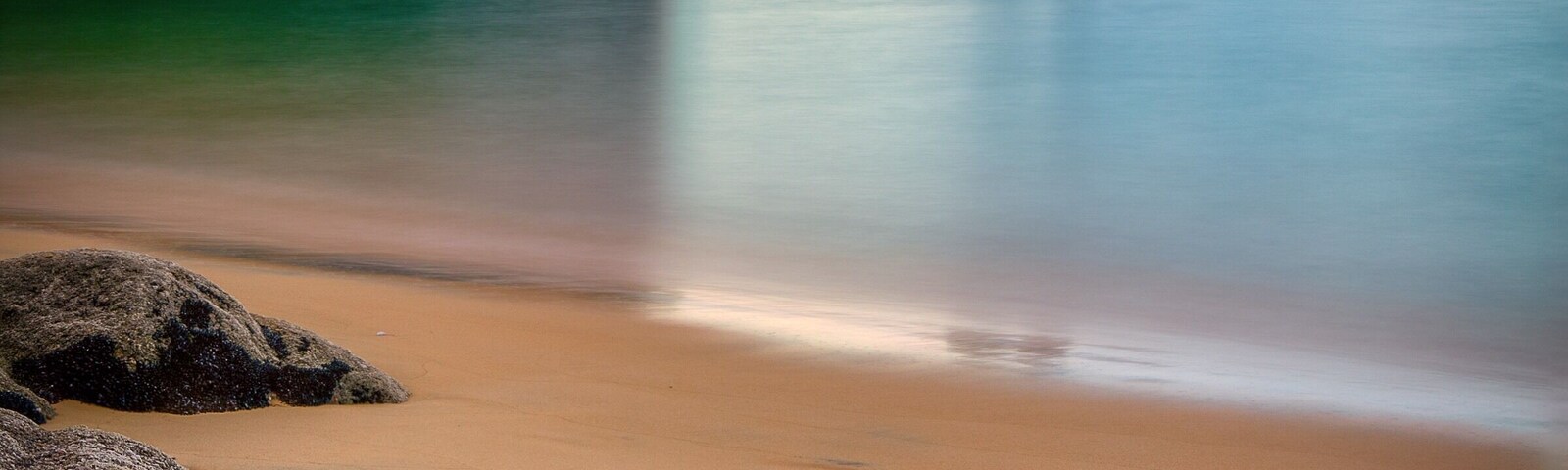 Uma praia com pedras grandes em primeiro plano e mar calmo.