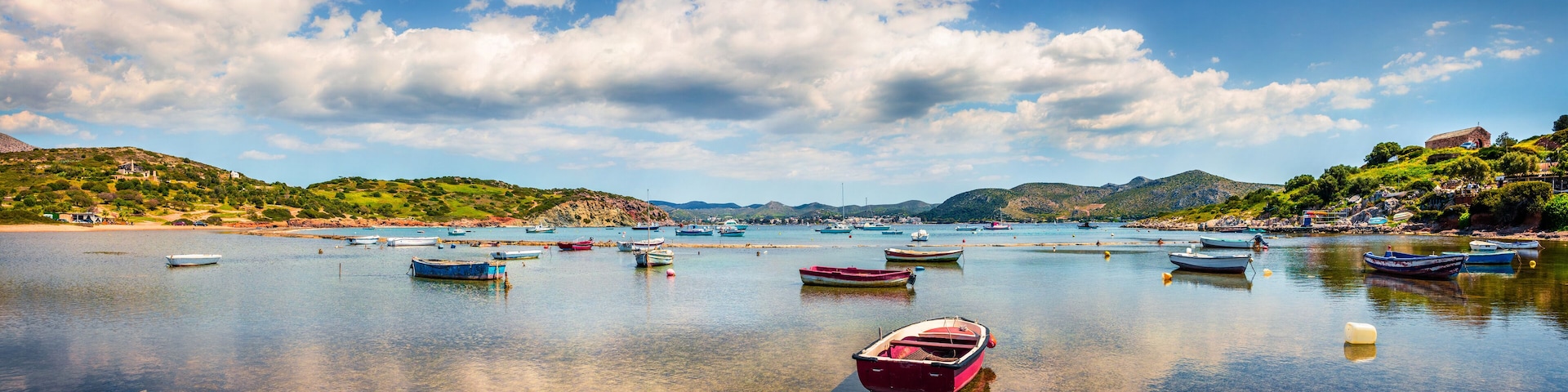 Uma baía calma com vários barcos ancorados, rodeada por colinas verdes e um céu azul e límpido.