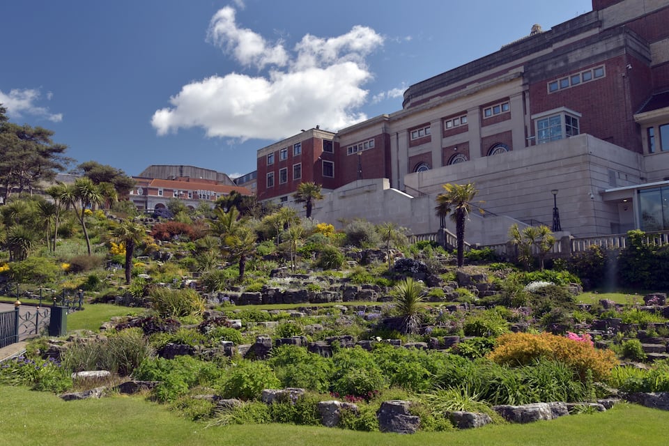 Um jardim ajardinado com diversas plantas e pedras, um edifício de tijolos vermelhos e um céu azul claro com nuvens.