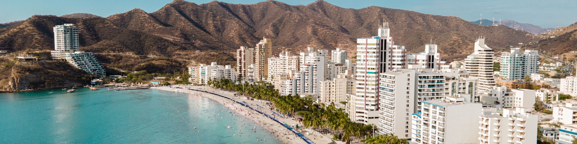Uma cidade costeira com praia, águas turquesa cristalinas e montanhas ao fundo.