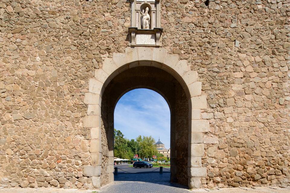 Um arco de pedra com uma estátua acima, dando acesso a uma estrada com carros e um edifício ao fundo.