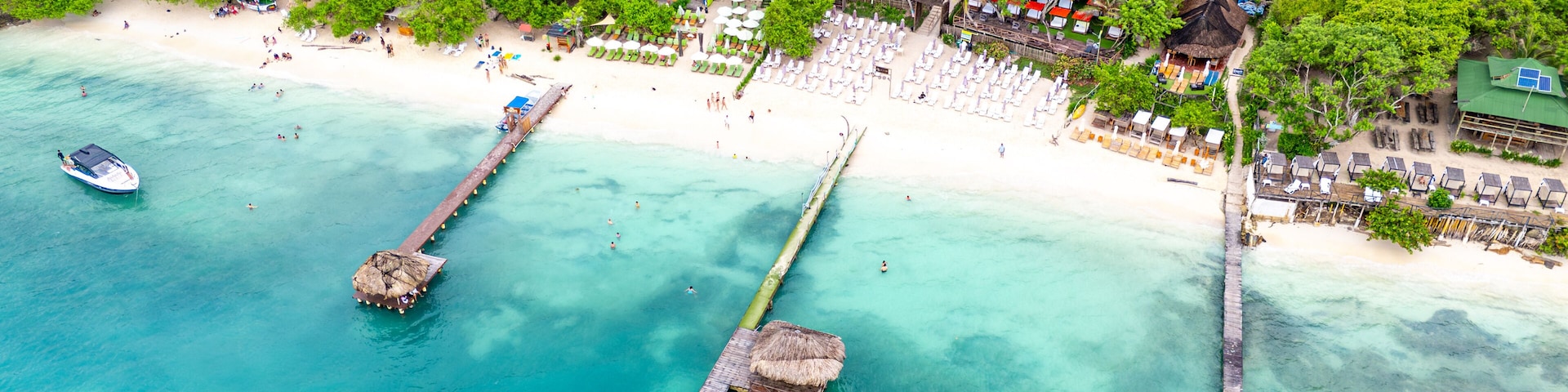 Uma praia tropical com águas turquesa cristalinas, um píer e uma área de resort com guarda-sóis e cadeiras de praia.