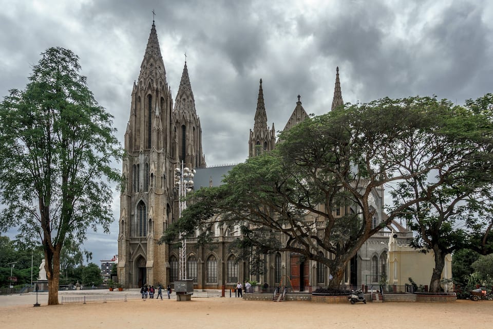 Uma catedral em estilo gótico, com diversas torres, cercada por árvores grandes e um calçadão.