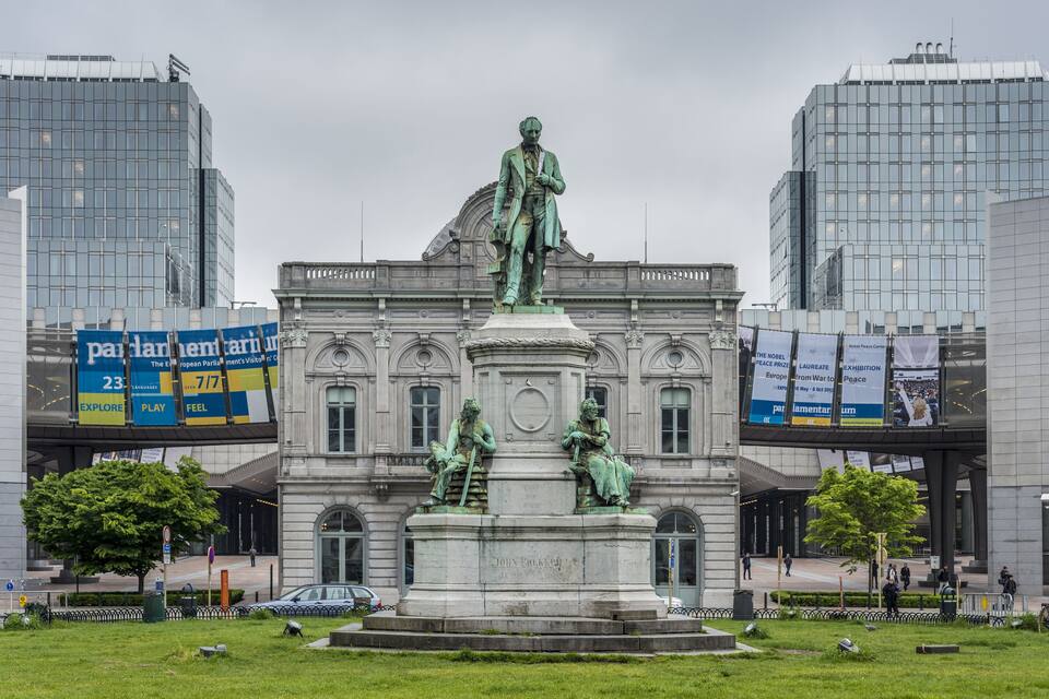 Estátua de um homem de terno, flanqueada por duas estátuas menores, em frente a um edifício com grandes faixas e uma estrutura de vidro moderna ao fundo.