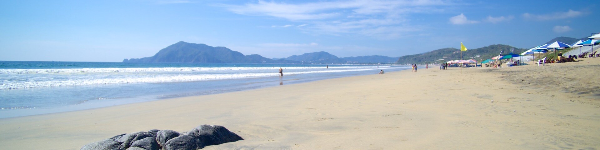 Uma praia arenosa com pedras grandes em primeiro plano, ondas quebrando na costa e montanhas ao longe.
