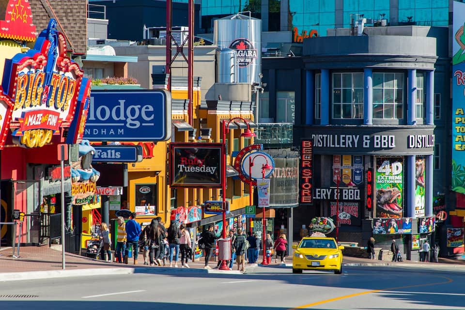 Uma rua vibrante com edifícios coloridos, incluindo uma placa 'Lodge Falls', uma placa 'Ruby Tuesday' e uma placa 'Distillery & BBQ'.