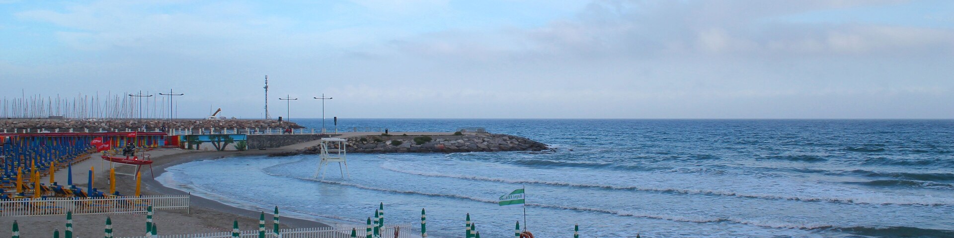 Uma praia com espreguiçadeiras e guarda-sóis verdes, um cais com barcos e um paredão rochoso.
