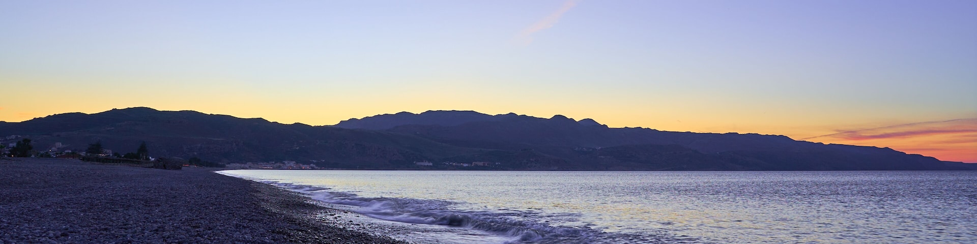Uma praia de pedras com mar calmo, montanhas ao longe e um céu ao pôr do sol.