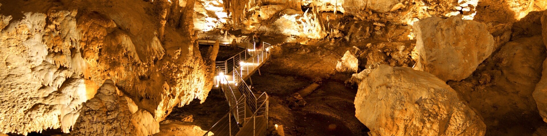 Caverna com estalactites e estalagmites, uma passarela metálica e uma grande formação rochosa.
