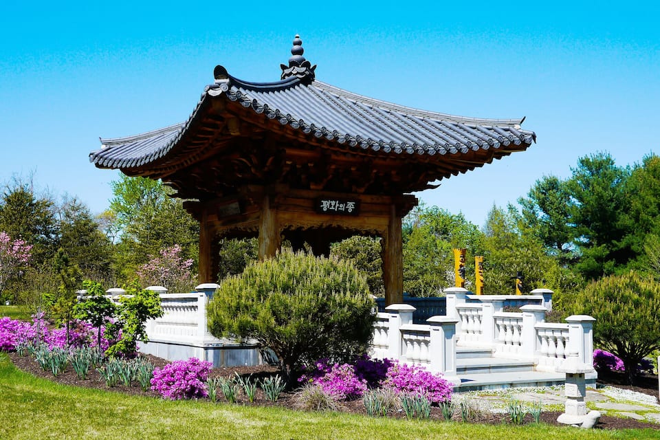 Um pavilhão tradicional coreano com telhado ladrilhado, rodeado por flores vibrantes e vegetação.