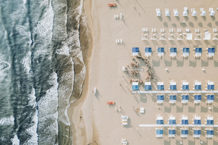 Vista aérea de uma praia com fileiras de espreguiçadeiras e algumas pessoas caminhando pela orla.