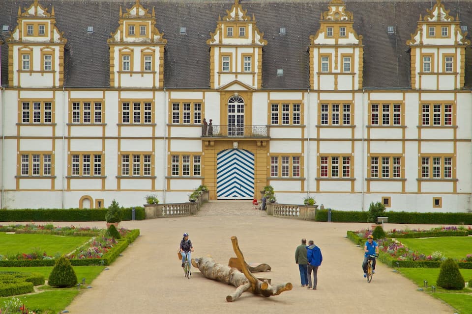 Um edifício grande e ornamentado com fachada simétrica, entrada central com um padrão azul e branco em chevron e um jardim bem cuidado com sebes e canteiros de flores.
