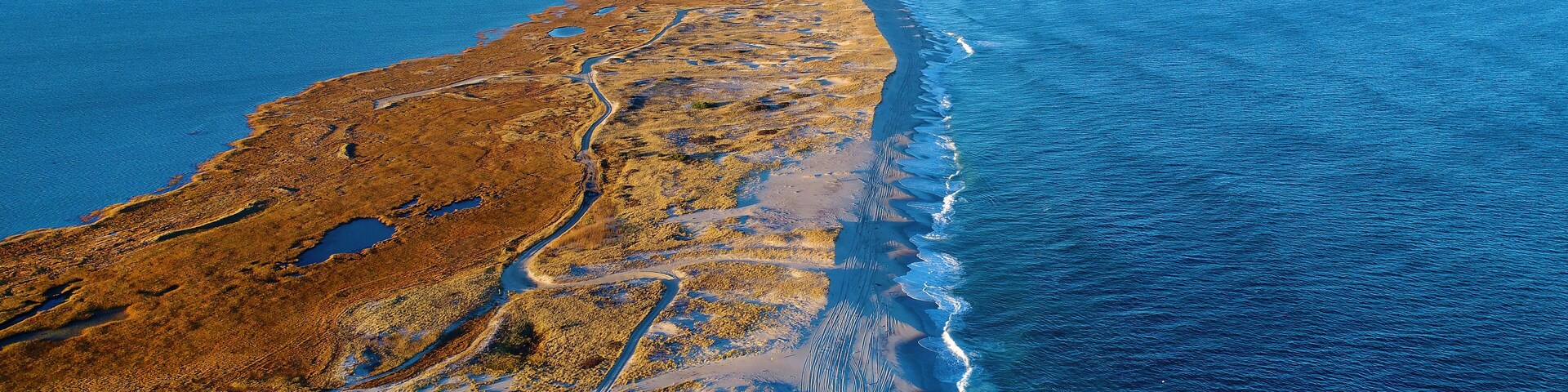 Paisagem costeira com uma praia arenosa, uma estrada sinuosa e uma grande extensão de água.