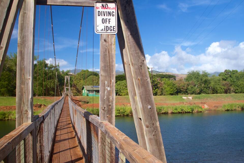 Uma ponte de madeira com uma placa indicando 'Não saltar da ponte', que leva a uma casa verde do outro lado do rio.