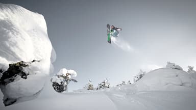 Um snowboarder executando um salto sobre uma paisagem coberta de neve, com árvores e grandes montes de neve.