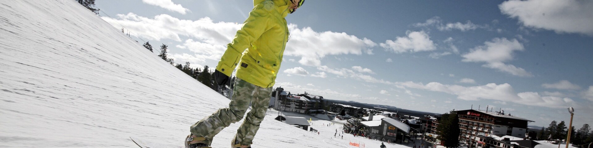 Um snowboarder com casaco e capacete amarelo desce uma encosta nevada, com edifícios e árvores ao fundo.